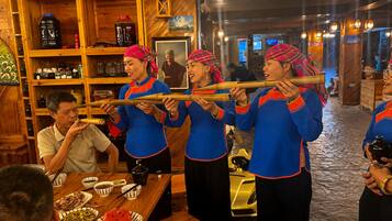 Um grupo de pessoas vestindo trajes típicos tocando um instrumento musical em um interior de madeira, com prateleiras e uma mesa com comida.