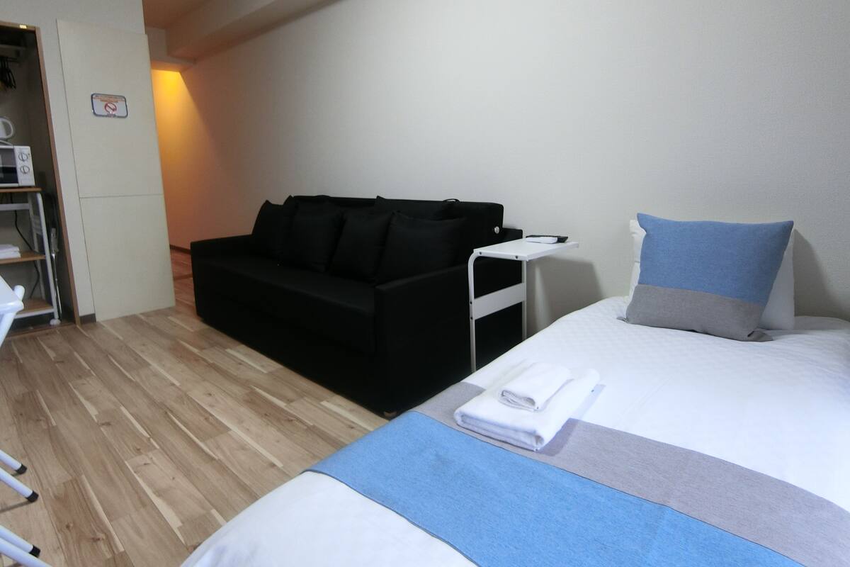 Quarto de hotel com uma cama, um sofá preto, uma mesinha e uma parede branca.