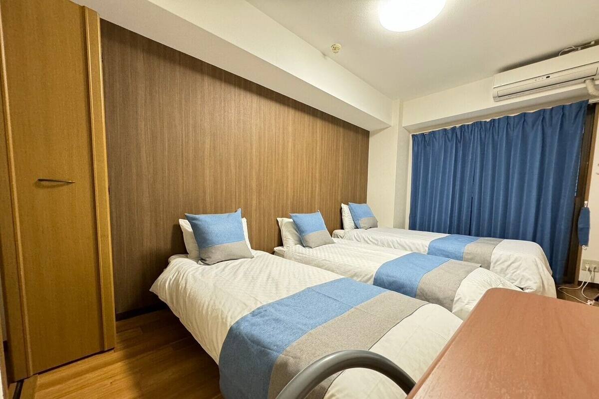 Quarto de hotel com duas camas, um guarda-roupa de madeira, uma escrivaninha e uma janela com cortinas.