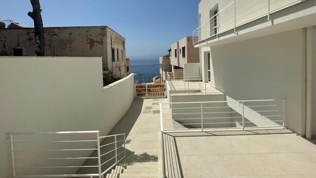 Corredor estreito e iluminado pelo sol, com vista para o mar, que leva a um edifício com varanda.