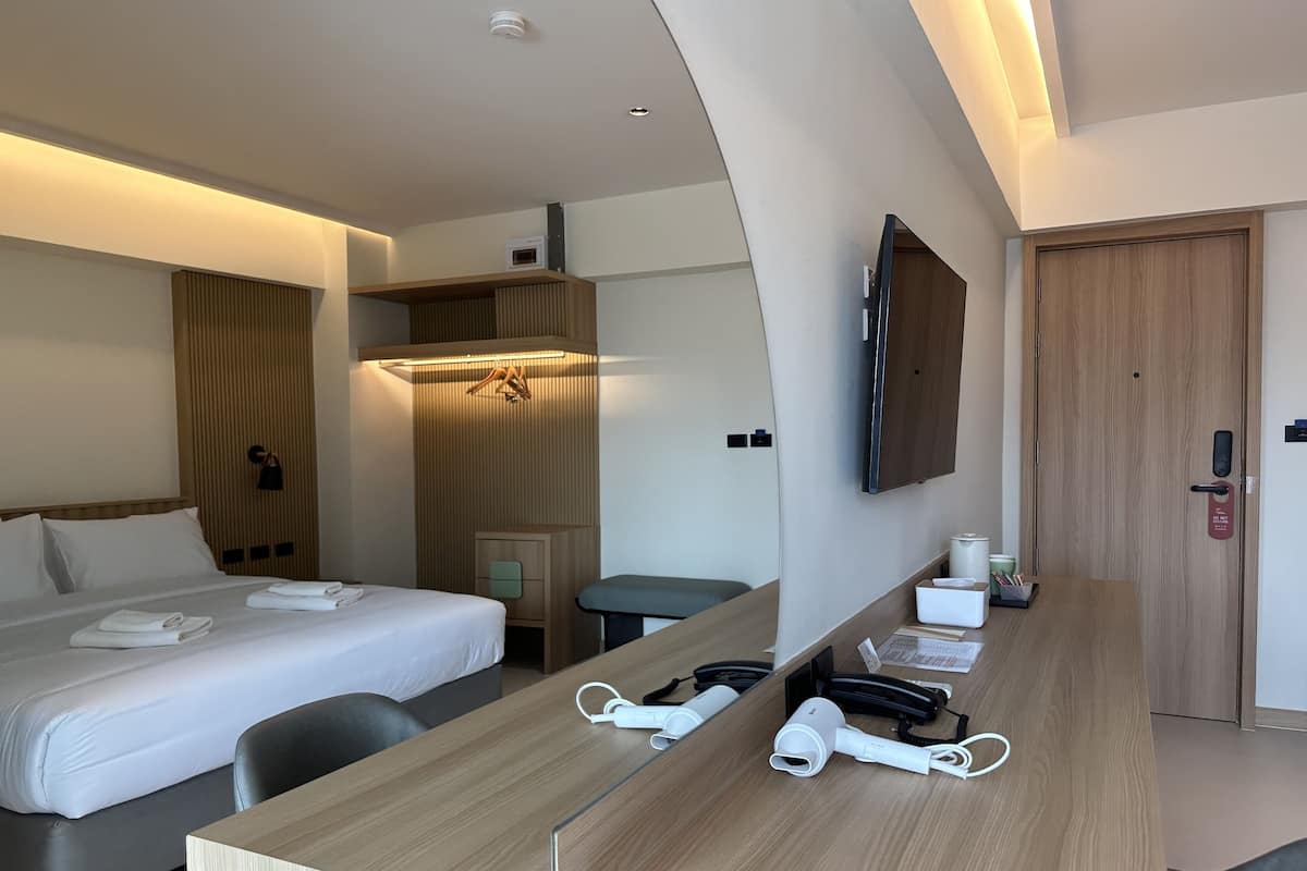 Quarto de hotel moderno com cama, mesa de cabeceira, escrivaninha e televisão fixada na parede.