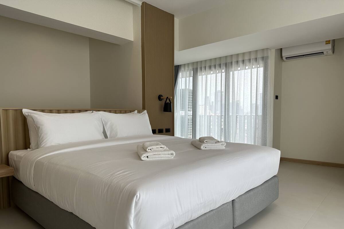 Quarto de hotel moderno com uma cama grande, uma mesinha de cabeceira, uma luminária e vista para a cidade através de cortinas translúcidas.