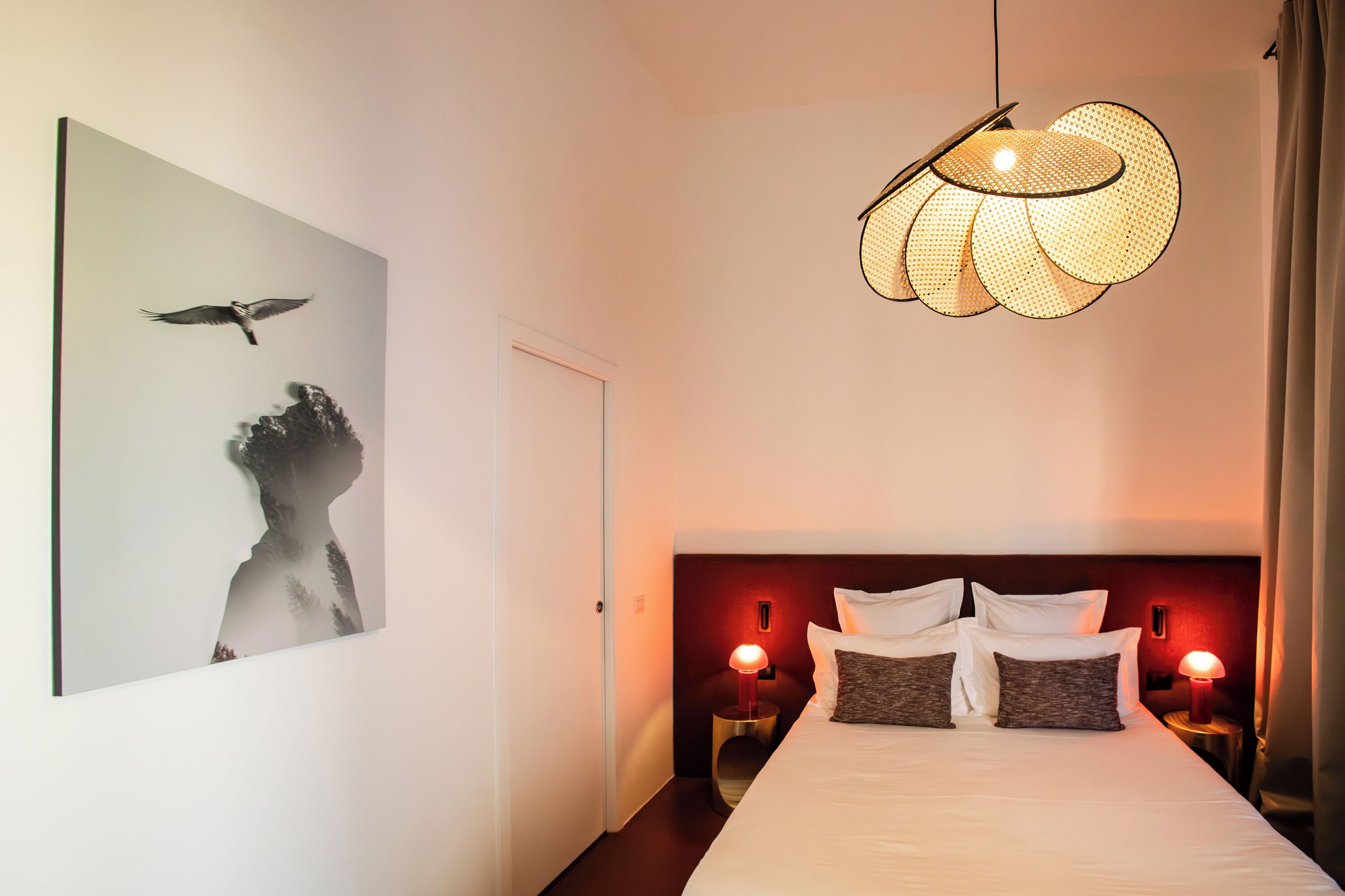 Quarto de hotel bem arrumado com uma cama grande, duas mesas de cabeceira com luminárias, uma obra de arte na parede retratando um pássaro e uma figura humana, e uma luminária pendente circular singular.