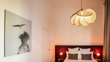 Quarto de hotel bem arrumado com uma cama grande, duas mesas de cabeceira com luminárias, uma obra de arte na parede retratando um pássaro e uma figura humana, e uma luminária pendente circular singular.