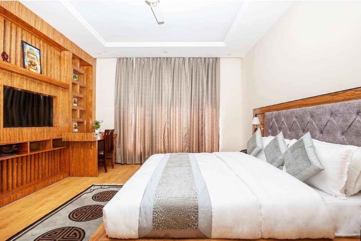 Quarto de hotel com cama, televisão fixada numa parede de madeira, escrivaninha e janela com cortinas.