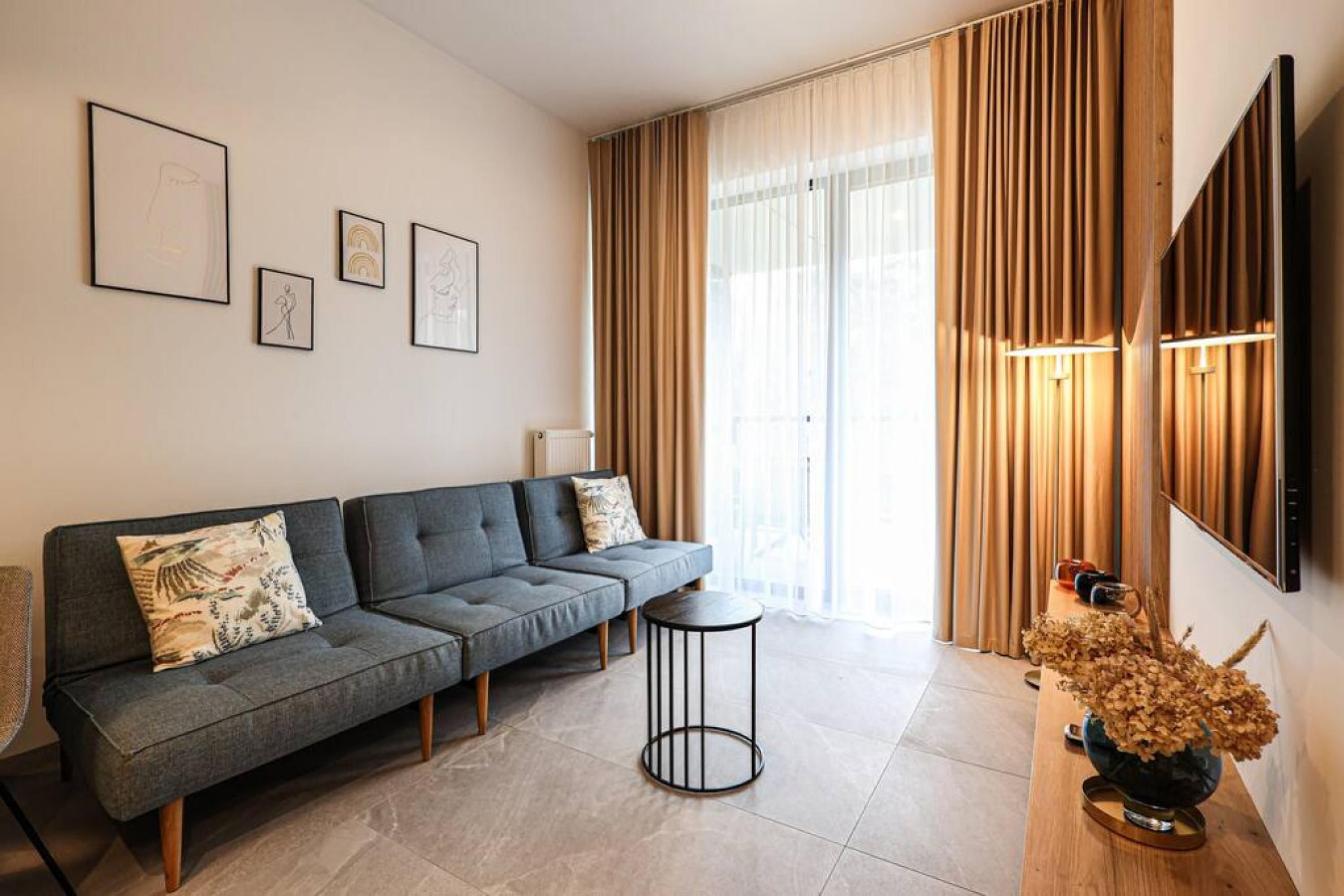 Uma sala de estar moderna com um sofá seccional cinza, uma mesa lateral redonda preta e um console de madeira com um vaso. Há obras de arte emolduradas na parede e uma janela grande com cortinas.