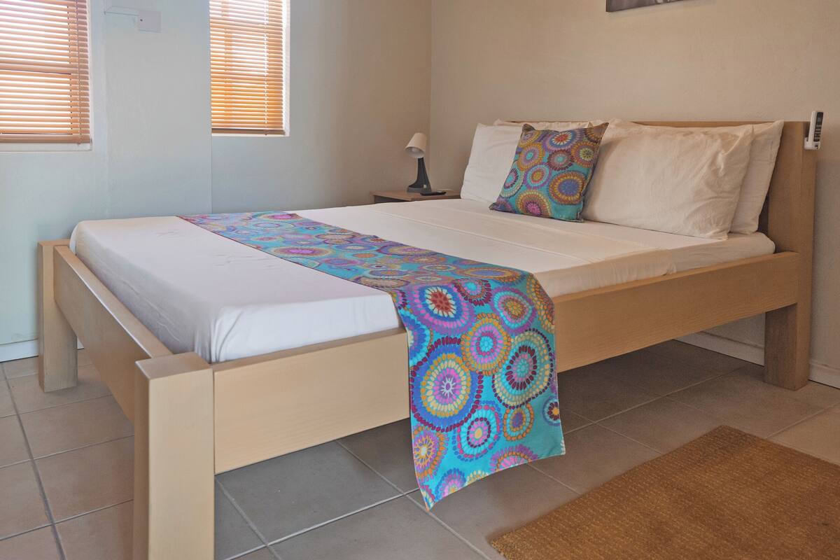 Quarto individual com cama de madeira, colcha colorida, duas almofadas, mesa de cabeceira com abajur e janela com persianas.