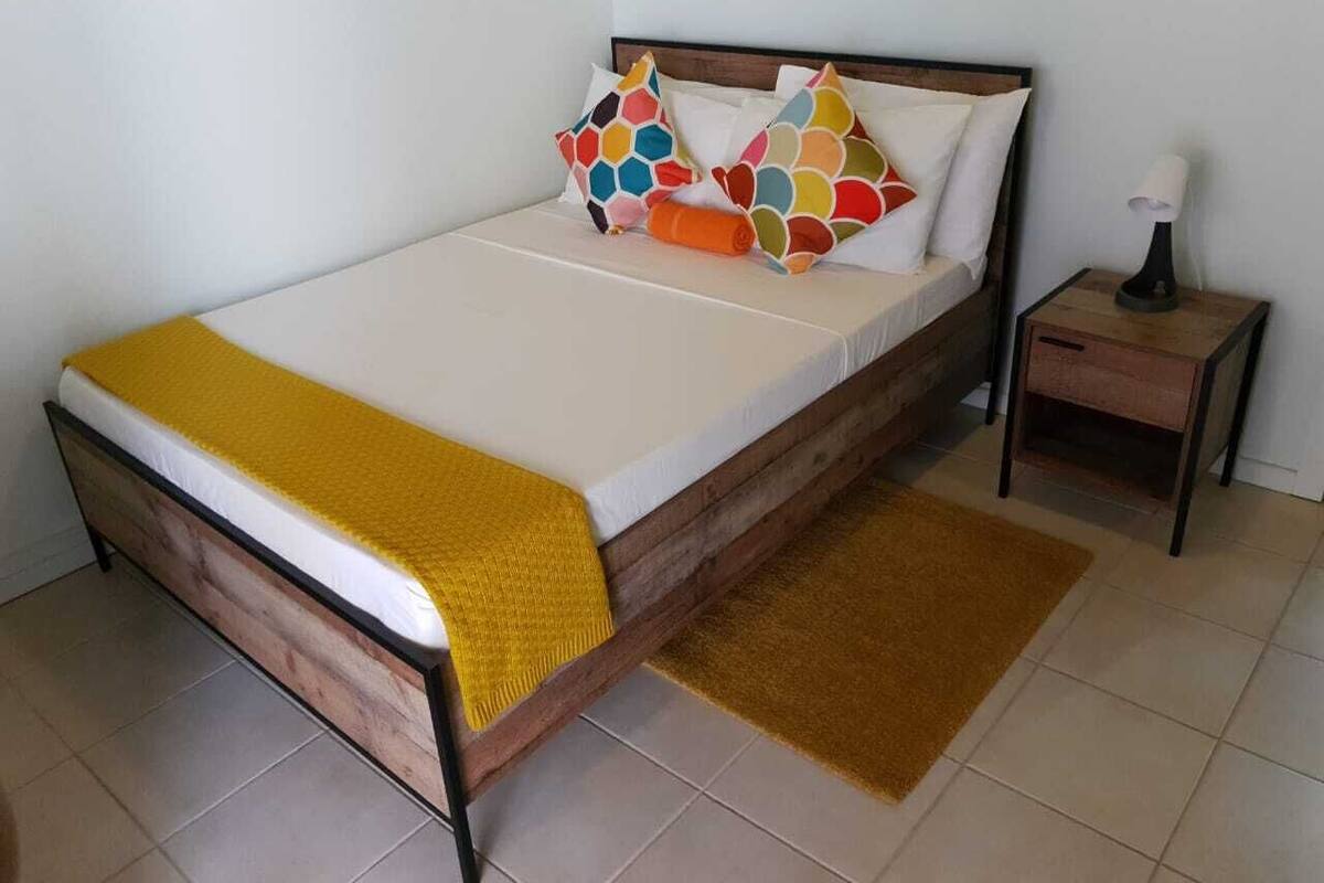 Cama com estrutura de madeira, roupa de cama branca e almofadas coloridas. Há uma pequena mesa de cabeceira com abajur, e um tapete marrom no chão.