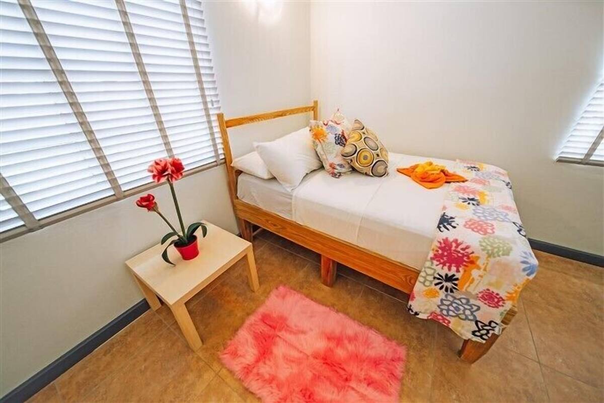 Quarto pequeno e bem organizado, com uma cama de solteiro, uma mesinha e um vaso de planta.