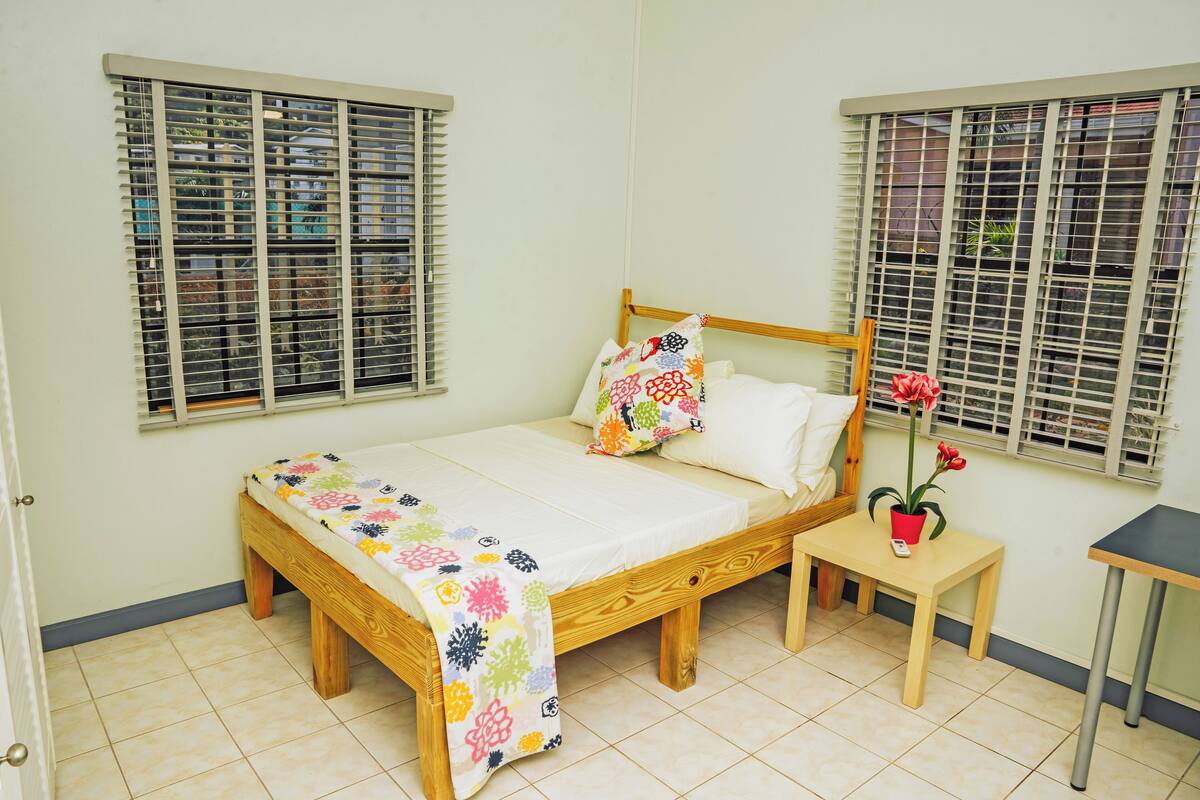 Quarto com cama de madeira, mesa de cabeceira com planta em vaso e duas janelas com persianas.
