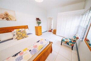 Quarto com cama de madeira, uma cadeira, cômoda e uma janela grande com persianas.