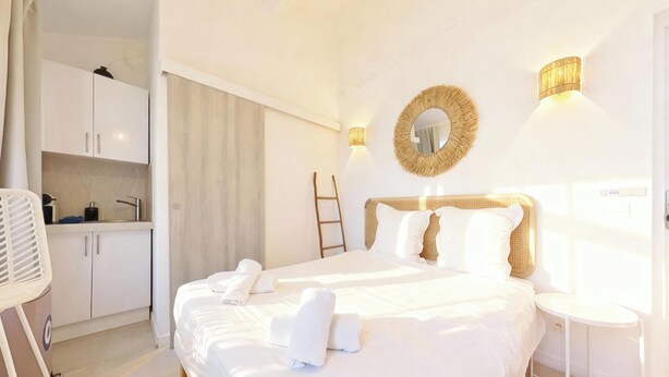 Quarto de hotel com cama, uma pequena mesa de cabeceira, espelho e área de cozinha compacta.