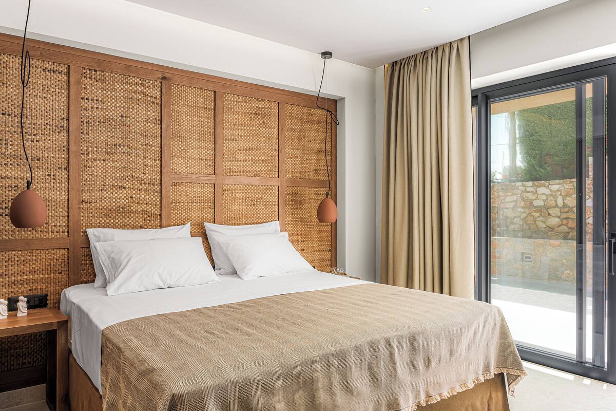 Quarto de hotel moderno com uma cama grande, cabeceira trançada, vista para o exterior e parede de pedra visível pela porta de vidro.