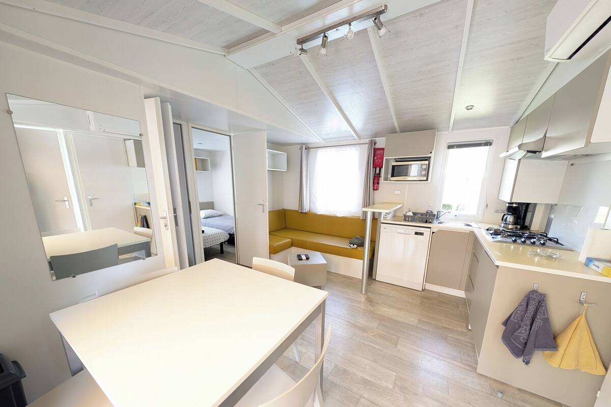 Quarto compacto e moderno, com mesa de jantar, sofá, pequena cozinha equipada com micro-ondas e fogão, e cama com vista para a janela.