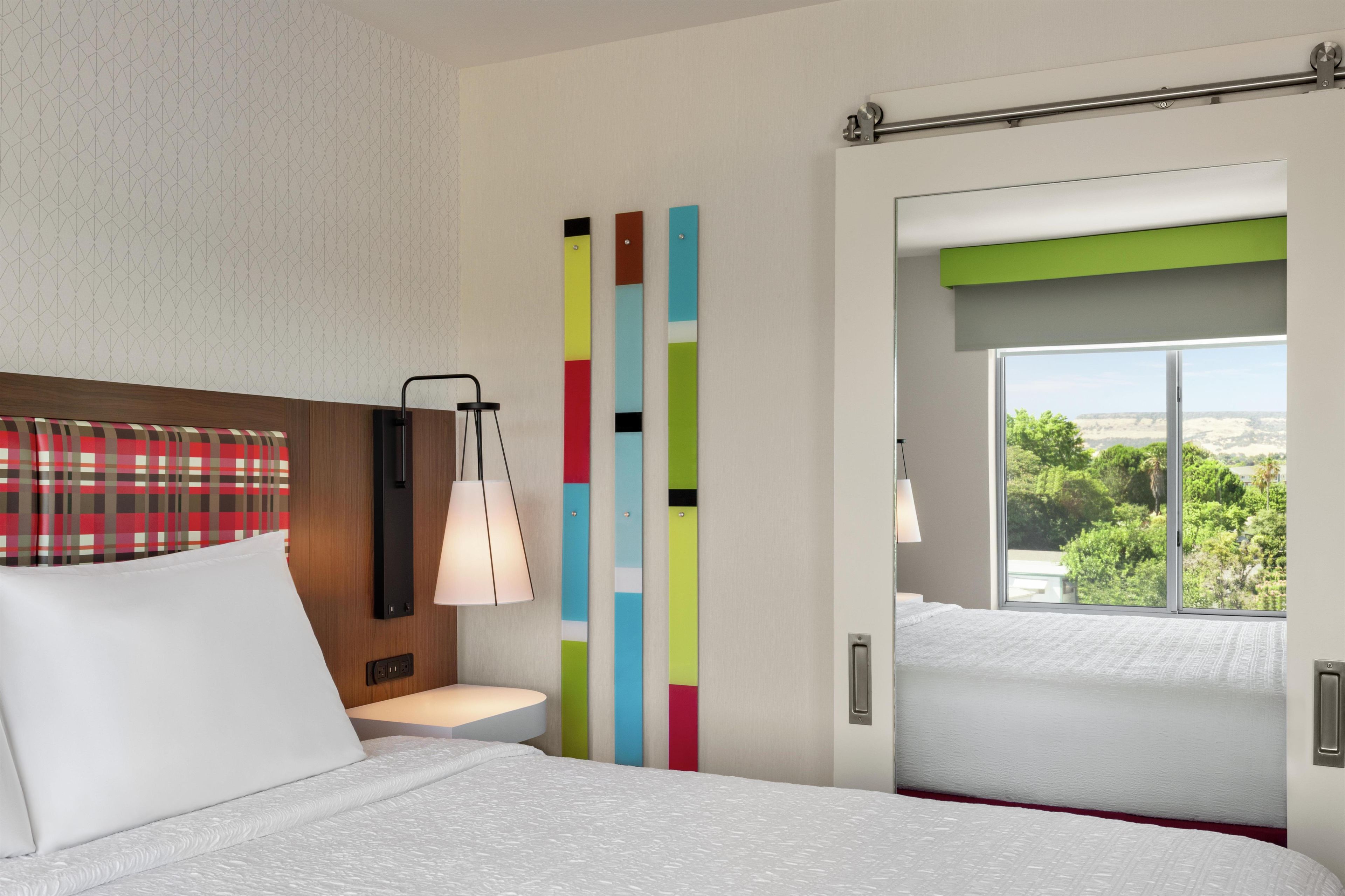 Quarto de hotel com cama, mesa de cabeceira, luminária, parede com listras verticais coloridas e janela com vista para áreas verdes.
