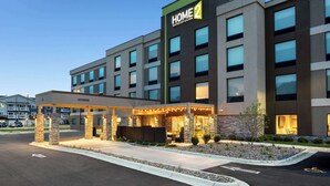 A modern hotel building with a sign reading "HOME 2 Suites by Hilton." It features a covered entrance with stone pillars and a well-lit outdoor area.
