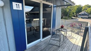 Um deck de madeira com uma porta de vidro deslizante marcada com o número 11, uma mesa pequena com cadeiras e um cercado com plantas em vasos.