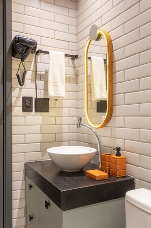 Casa de banho com lavatório branco, espelho com moldura dourada, porta-toalhas com duas toalhas e secador de cabelo preso à parede.