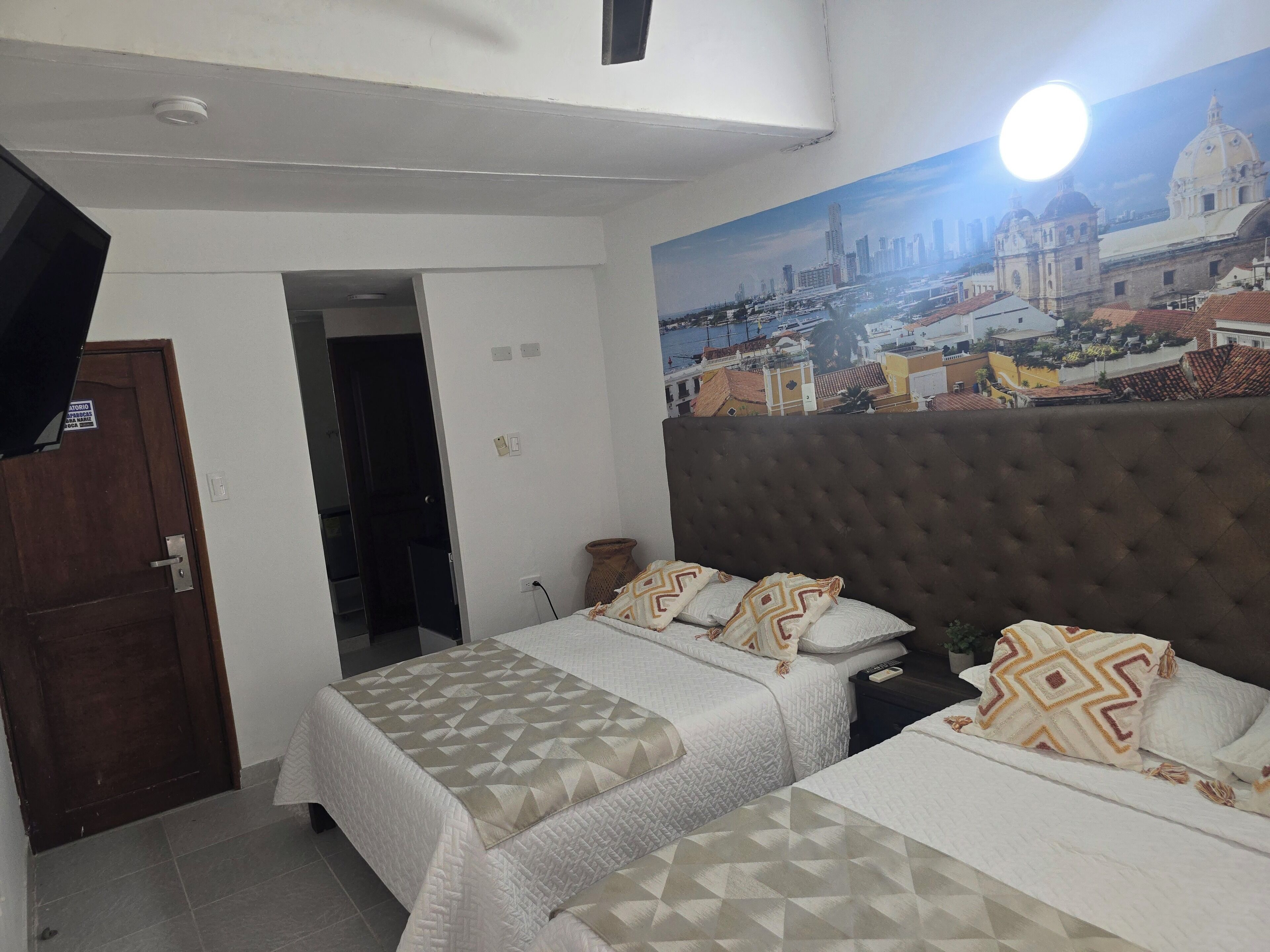 Quarto de hotel com duas camas, um grande mural de uma paisagem urbana, cabeceira marrom e uma porta à esquerda.