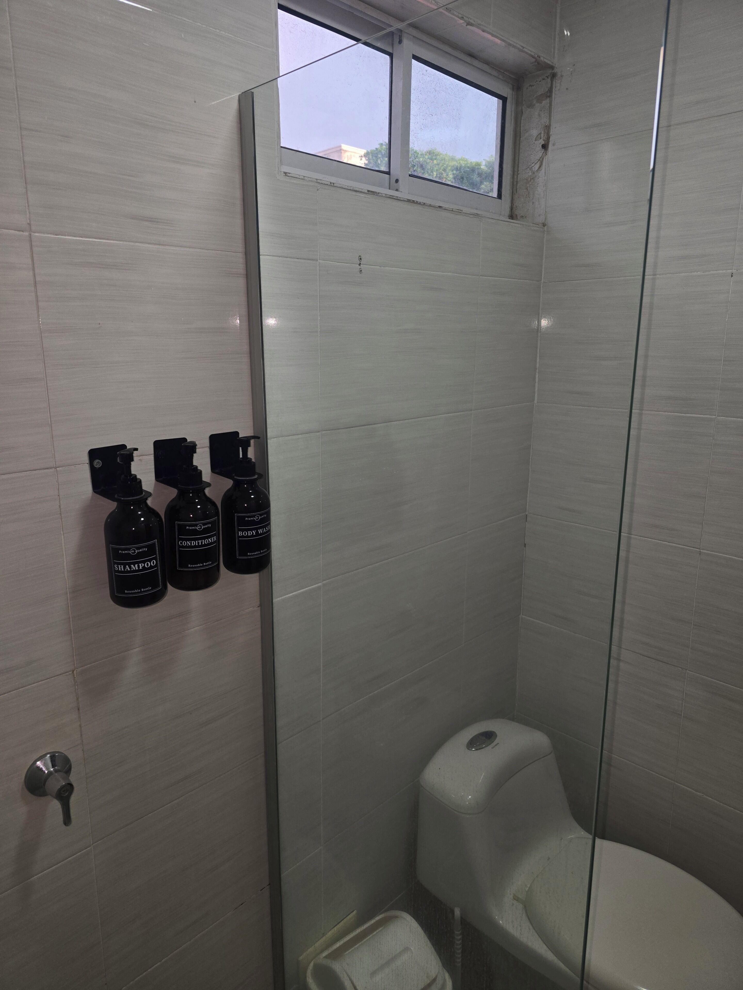 Box de chuveiro com porta de vidro, vaso sanitário branco e três frascos pretos de shampoo e condicionador na parede.