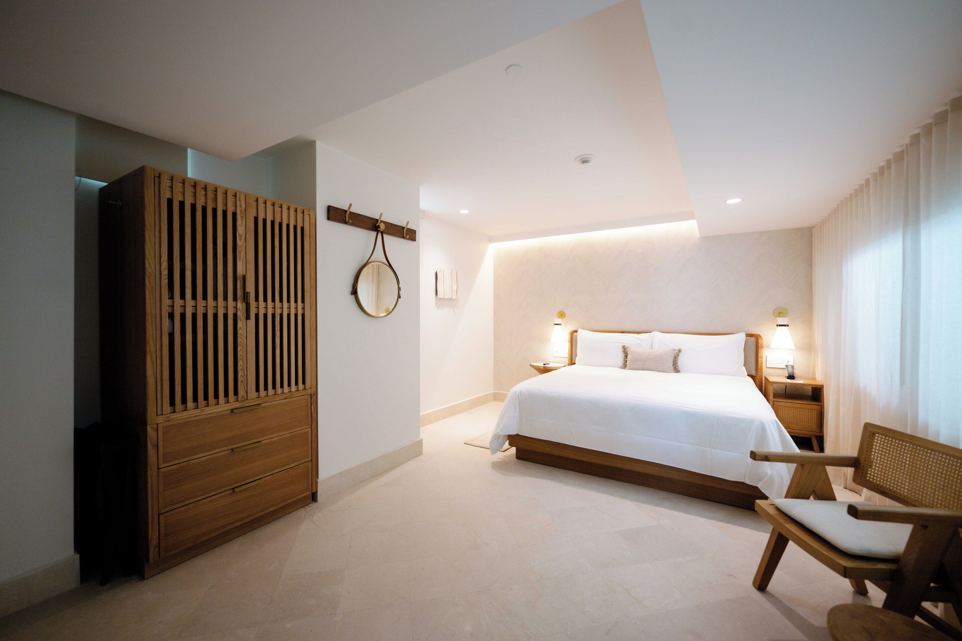 Quarto de hotel com cama, guarda-roupa de madeira, cadeira, mesa de cabeceira com abajur e espelho fixado na parede.