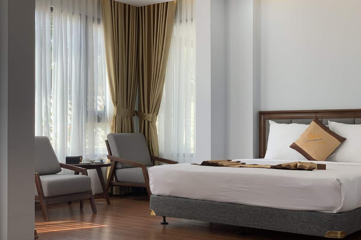 Quarto de hotel com cama, duas cadeiras, uma mesinha e cortinas leves.