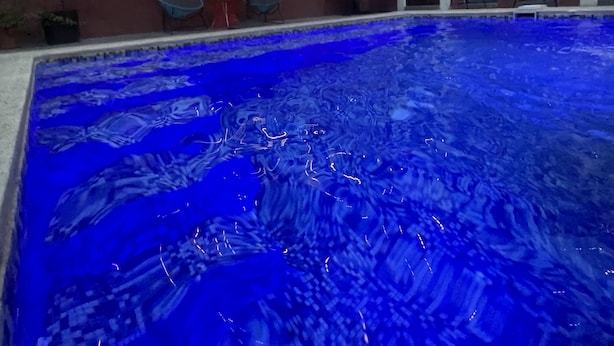 Piscina com design em mosaico azul, rodeada por uma parede vermelha com uma cadeira suspensa e um pequeno guarda-sol.