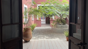 Porta aberta para um pátio com azulejos estampados, plantas em vasos e um edifício vermelho ao fundo.