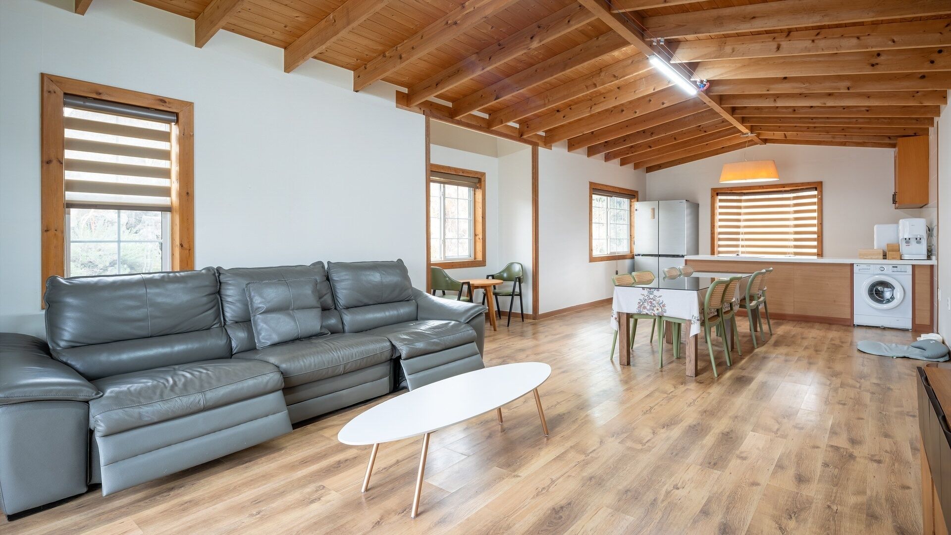 Um espaço de estar moderno com sofá de couro, uma pequena mesa branca, piso de madeira e uma área de cozinha com máquina de lavar.
