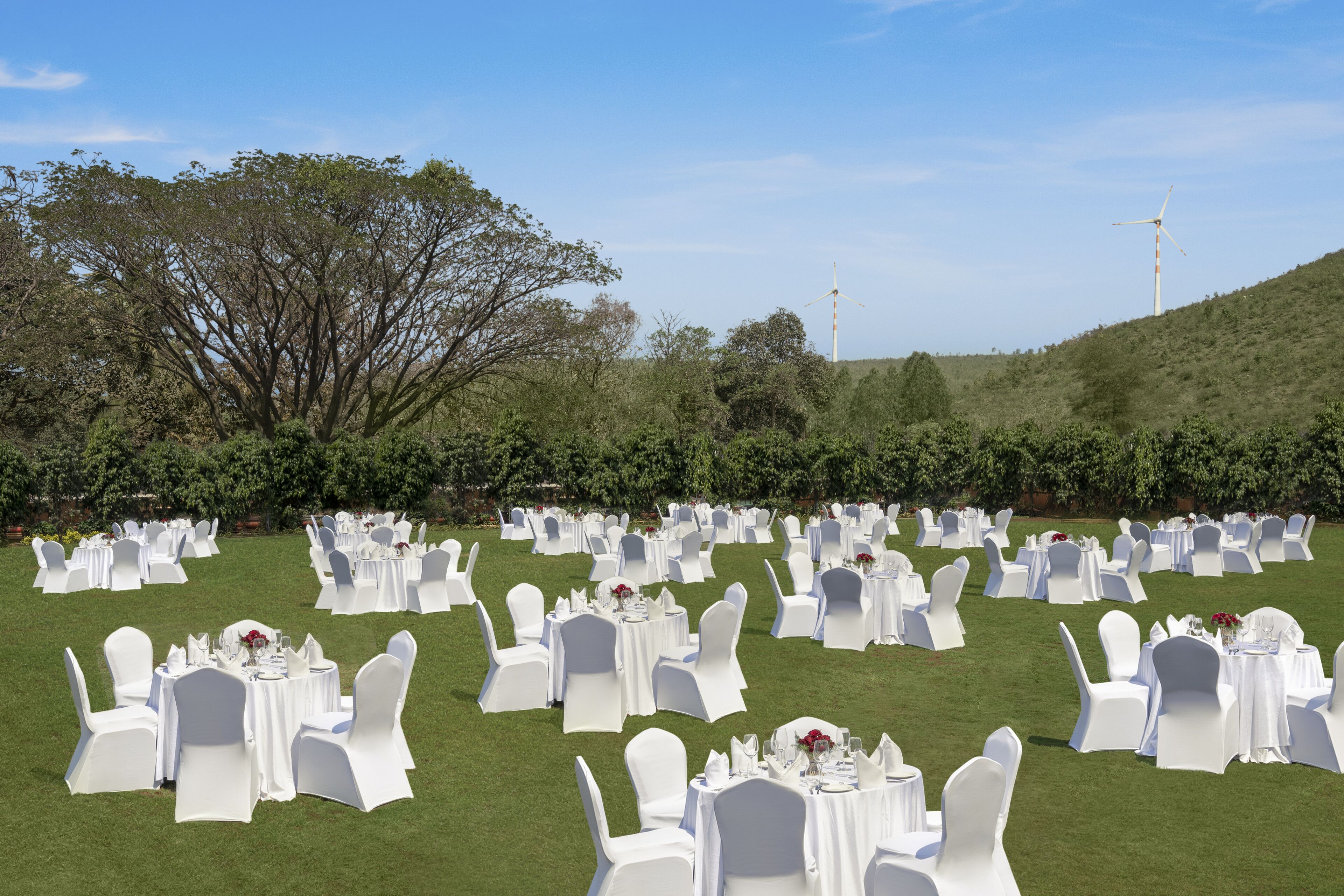 Espaço para casamento ao ar livre com fileiras de mesas e cadeiras brancas, com vista para árvores e uma colina.