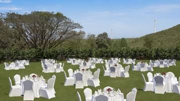 Espaço para casamento ao ar livre com fileiras de mesas e cadeiras brancas, com vista para árvores e uma colina.
