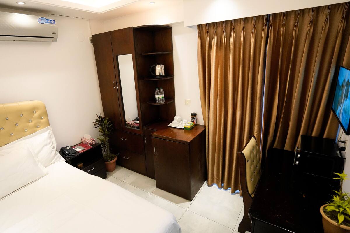 Quarto de hotel com cama, escrivaninha, guarda-roupa, televisão e um vaso de planta.