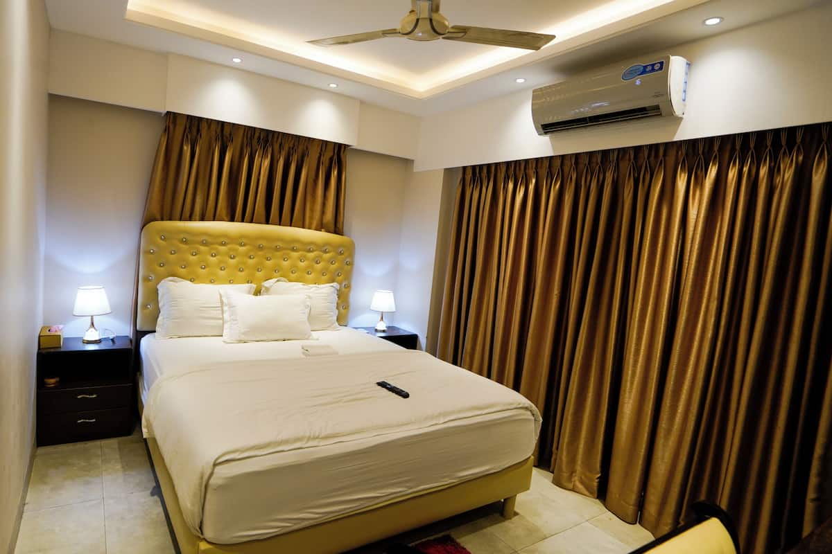 Quarto de hotel com uma cama grande, duas mesas de cabeceira com luminárias, um ventilador de teto e um aparelho de ar condicionado.