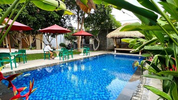 Uma piscina com borda revestida, rodeada por cadeiras verdes e guarda-sóis vermelhos, e uma estrutura de telhado de palha ao fundo.