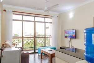 Uma sala de estar ampla com uma janela grande, ventilador de teto, televisão de tela plana e um dispenser de água.