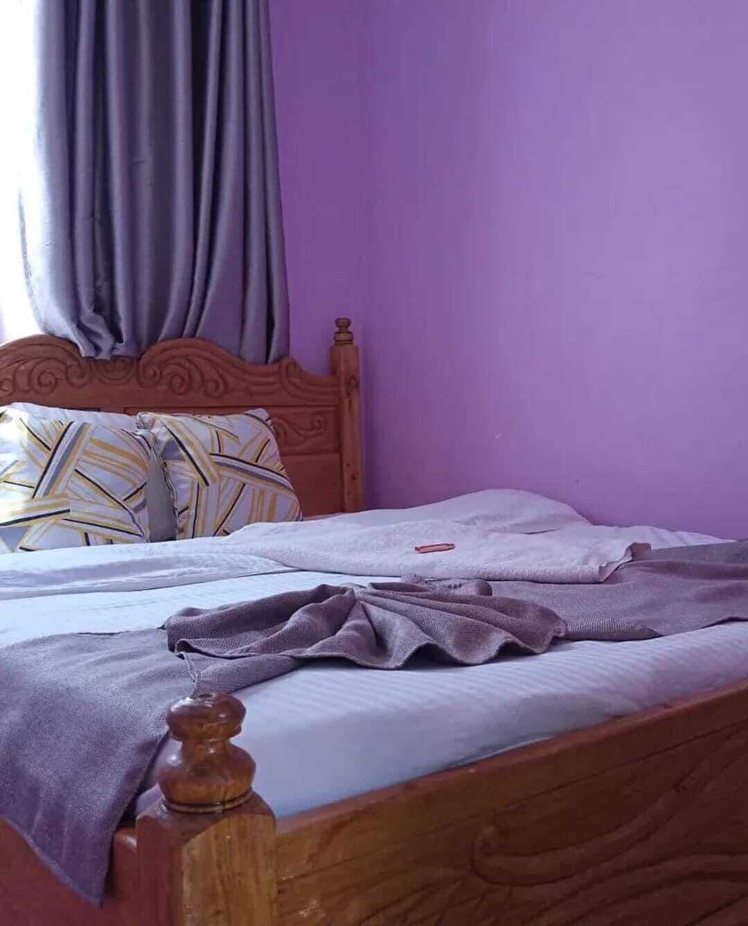 Uma cama com cabeceira de madeira, parede roxa e janela com cortina.