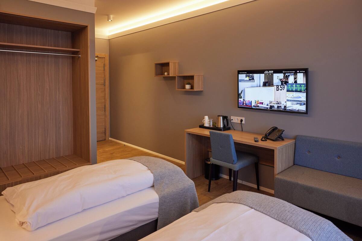 Quarto de hotel com duas camas, uma escrivaninha com cadeira, um sofá, uma televisão e uma pequena estante com uma planta.