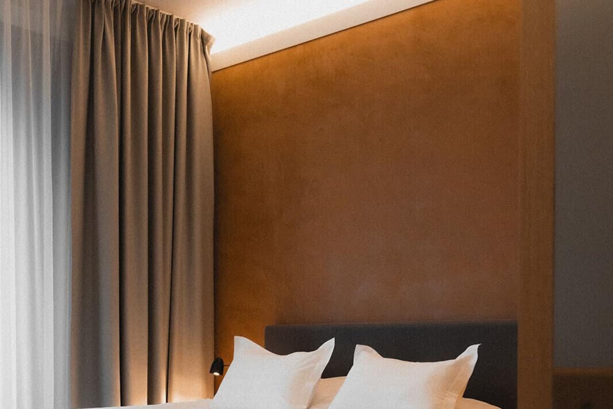 Quarto de hotel moderno com cabeceira de madeira, cama grande com roupa de cama branca e manta bege.