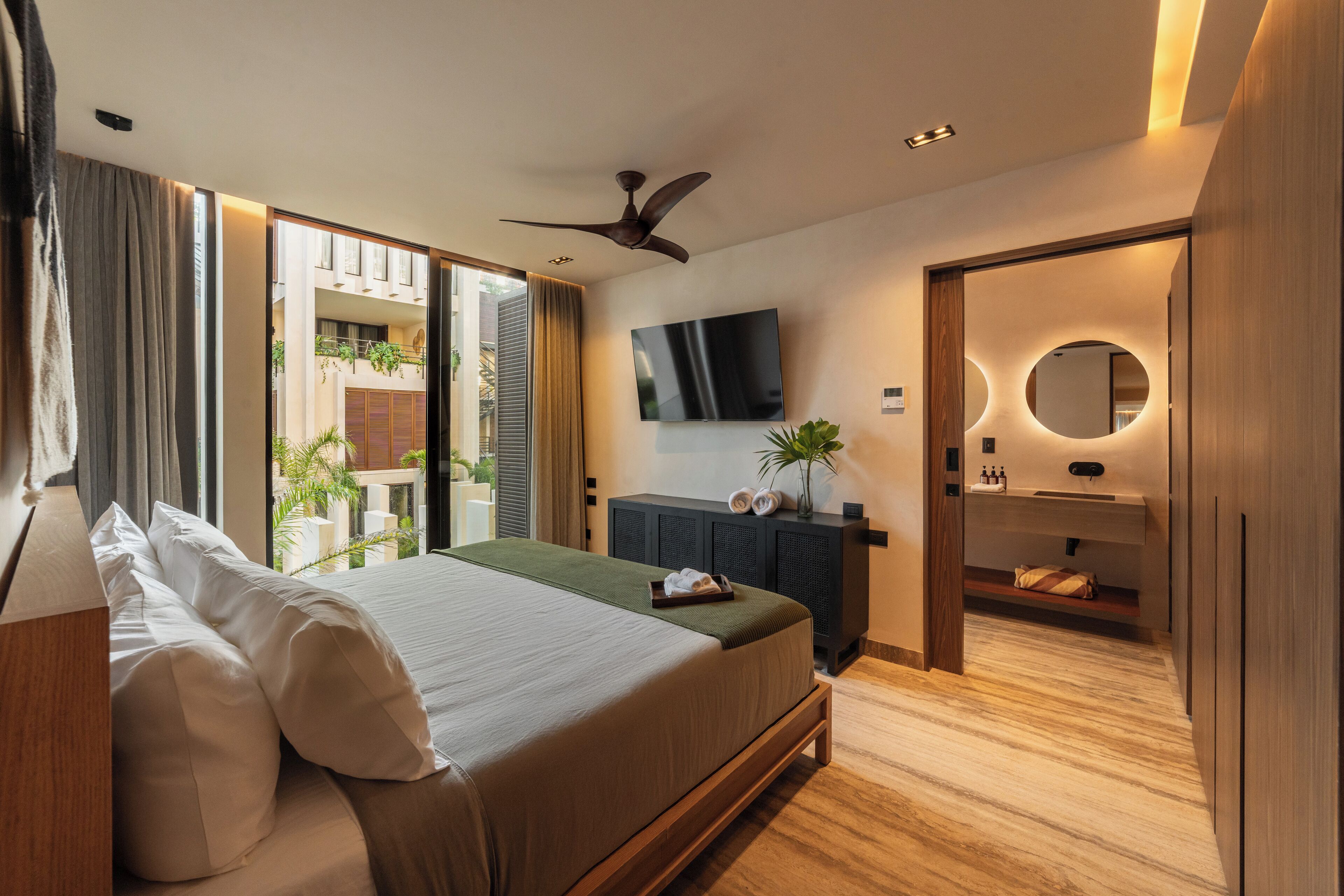 Quarto de hotel moderno com uma cama grande, ventoinha no teto, televisão, escrivaninha pequena e banheiro com espelho e pia.