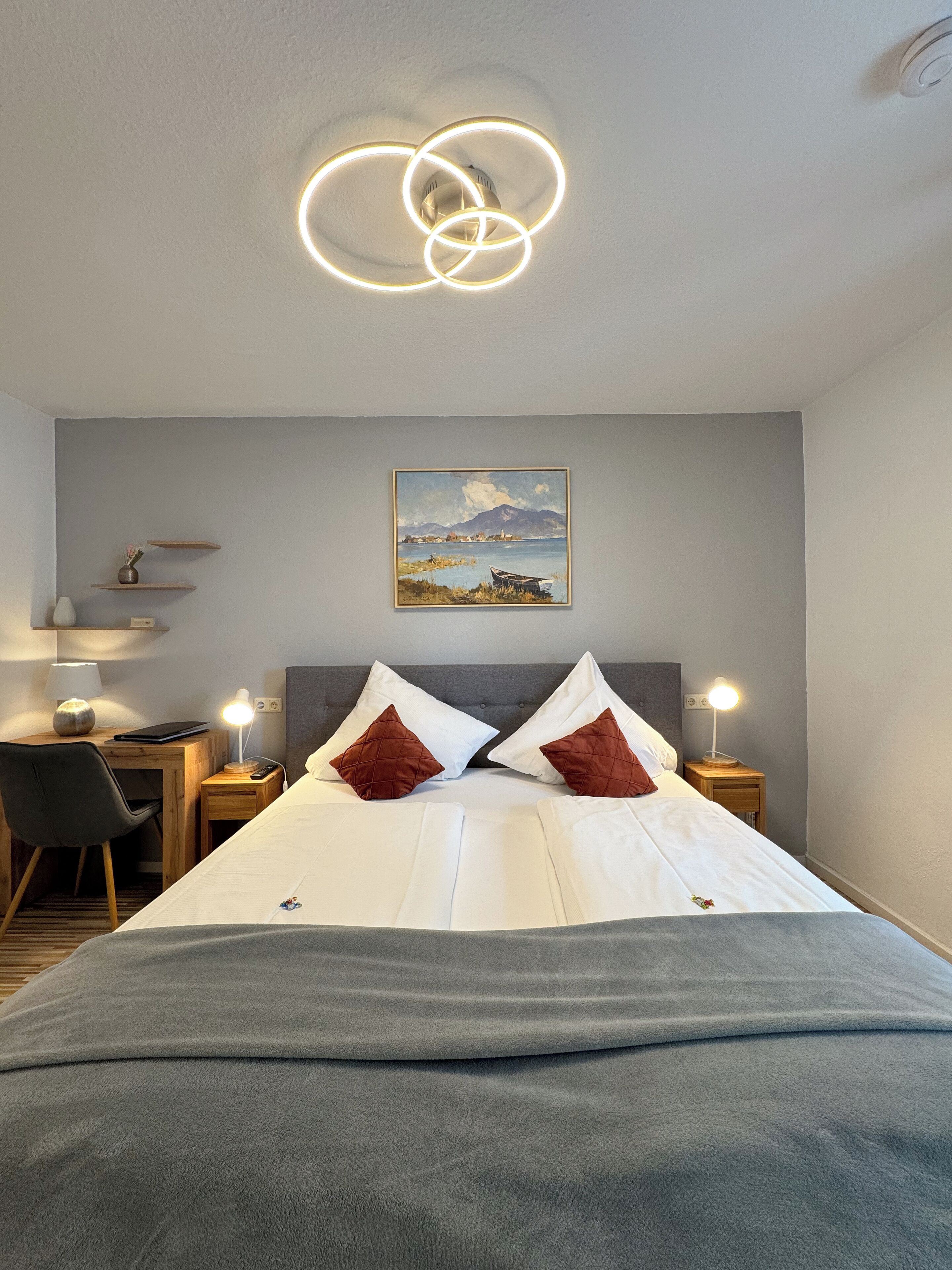Quarto de hotel moderno com uma cama bem arrumada, uma escrivaninha com abajur, uma cadeira e um quadro de uma paisagem na parede.