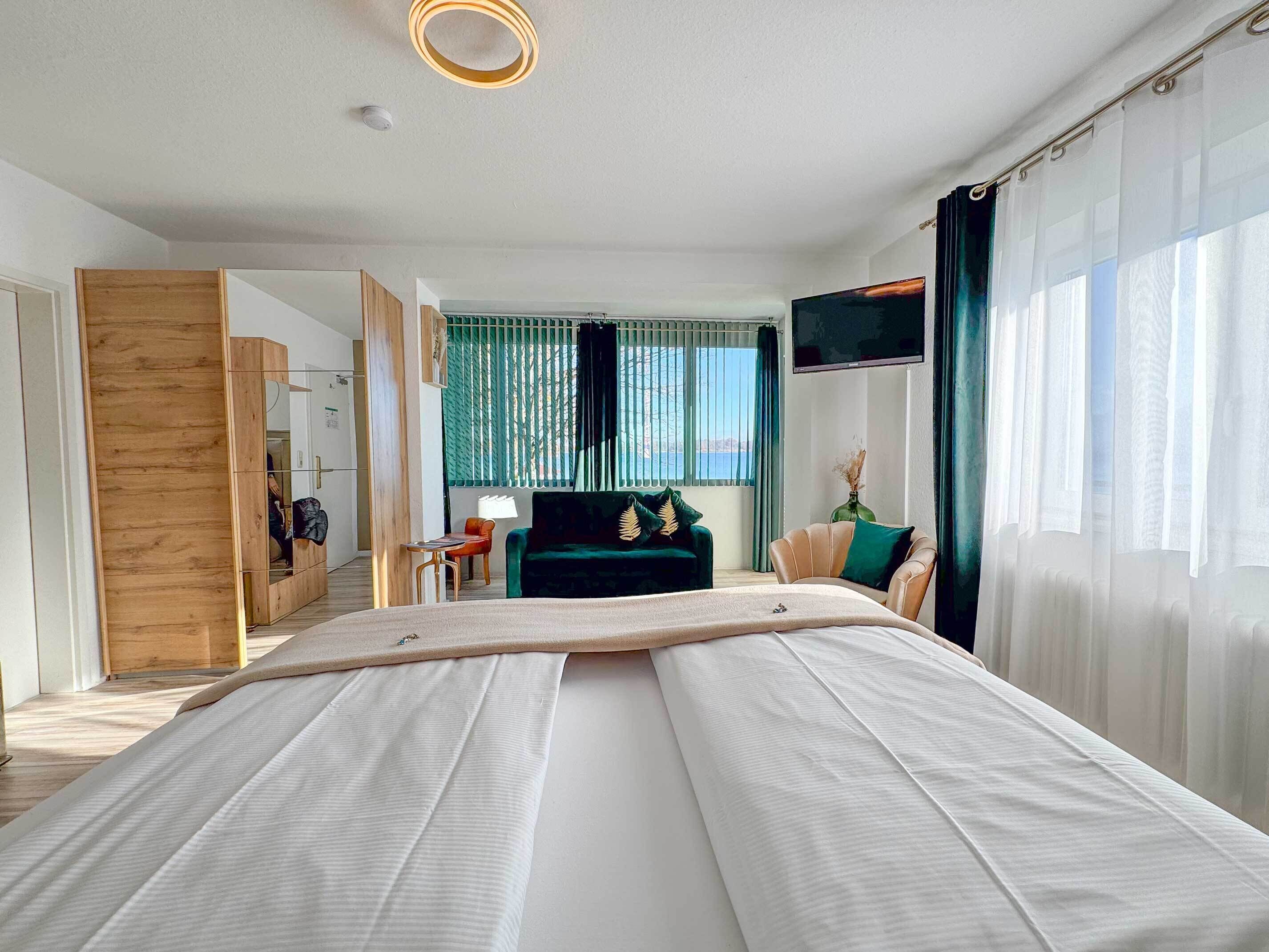 Quarto de hotel moderno com uma cama grande, televisão presa à parede e vista para o mar pelas janelas.