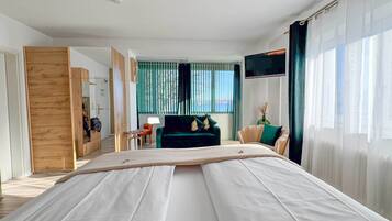 Quarto de hotel moderno com uma cama grande, televisão presa à parede e vista para o mar pelas janelas.