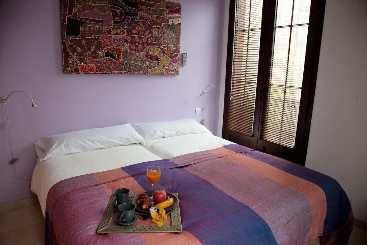 Quarto de hotel com cama, manta colorida, bandeja com frutas e bebida, e uma grande pintura abstrata na parede.