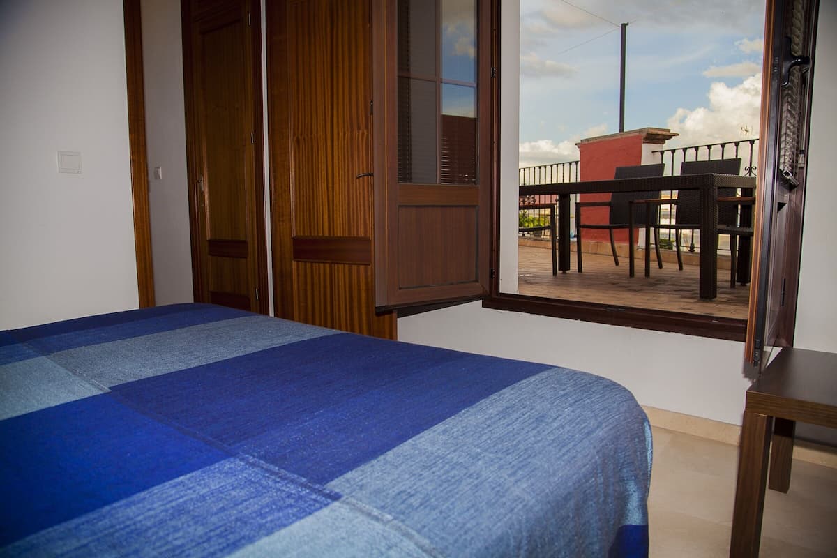Quarto com uma cama, porta de madeira e vista para uma varanda externa com mesa e cadeiras.
