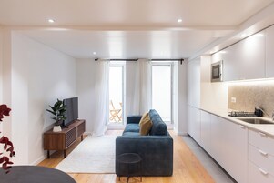 Um apartamento moderno com sala de estar, incluindo sofá, TV e uma pequena mesinha de apoio. Possui cozinha com armários brancos e micro-ondas.