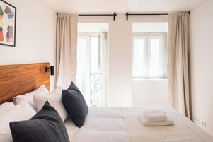 Cama bem arrumada com almofadas brancas e escuras, cabeceira de madeira e janela com cortinas.