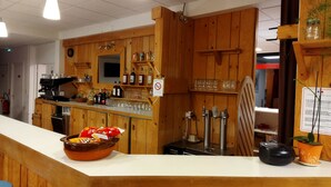 Uma área de bar com balcão de madeira, máquina de café, garrafas, copos e uma tigela de petiscos.