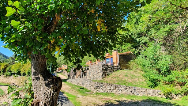 Um edifício de paredes de pedra com telhado vermelho, rodeado por vegetação exuberante e um céu azul límpido.