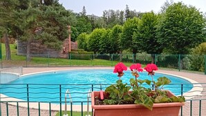 Uma piscina cercada por uma cerca verde, com um vaso de flores cor-de-rosa em primeiro plano.