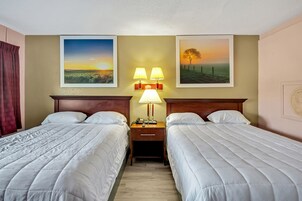 Quarto de hotel com duas camas de solteiro, uma mesa de cabeceira com abajur e duas fotos emolduradas na parede.